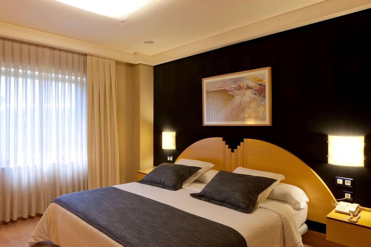 Quarto de hotel com uma cama grande, duas mesas de cabeceira, um quadro na parede e uma janela com cortinas.