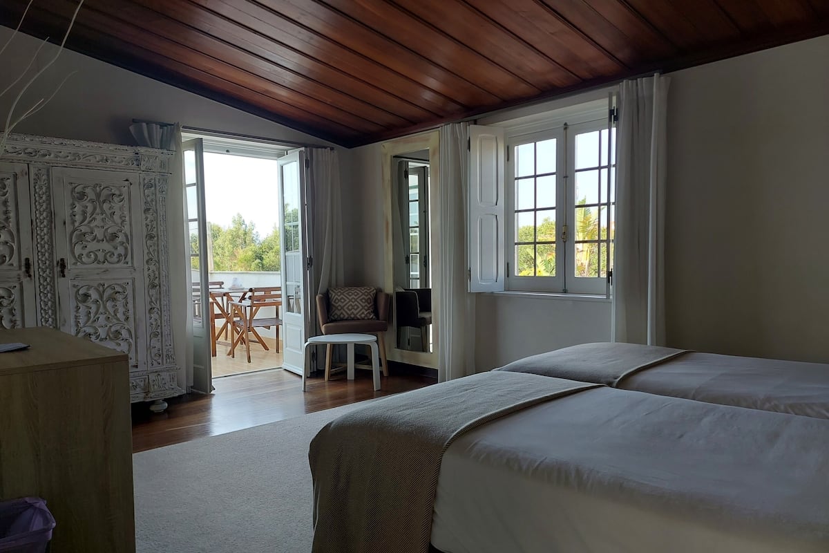 Quarto com duas camas, teto de madeira, guarda-roupa, uma mesinha e vista para o exterior.