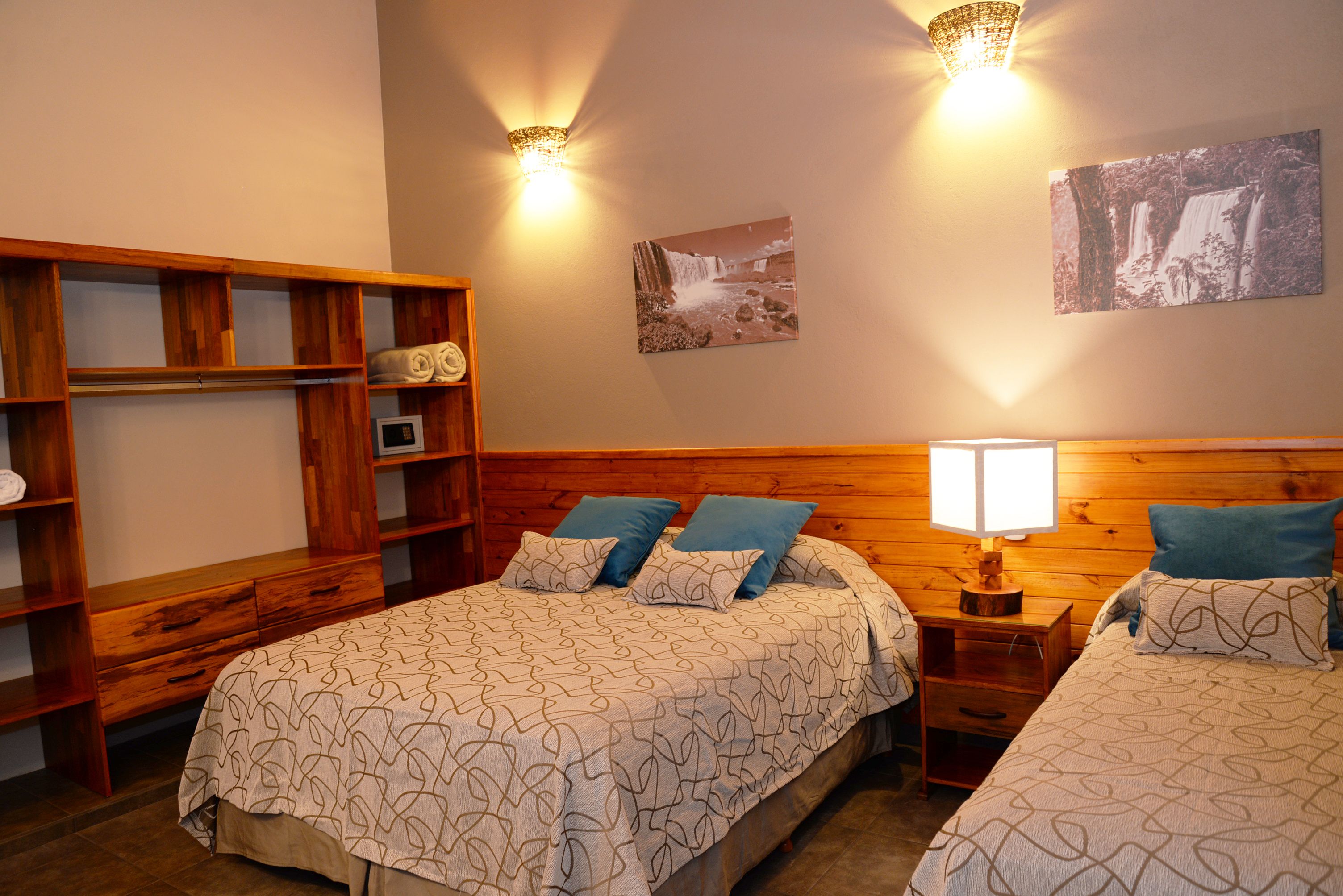 Quarto de hotel com duas camas, cabeceira de madeira, prateleiras fixadas na parede, mesa de cabeceira com abajur e duas luzes de parede.