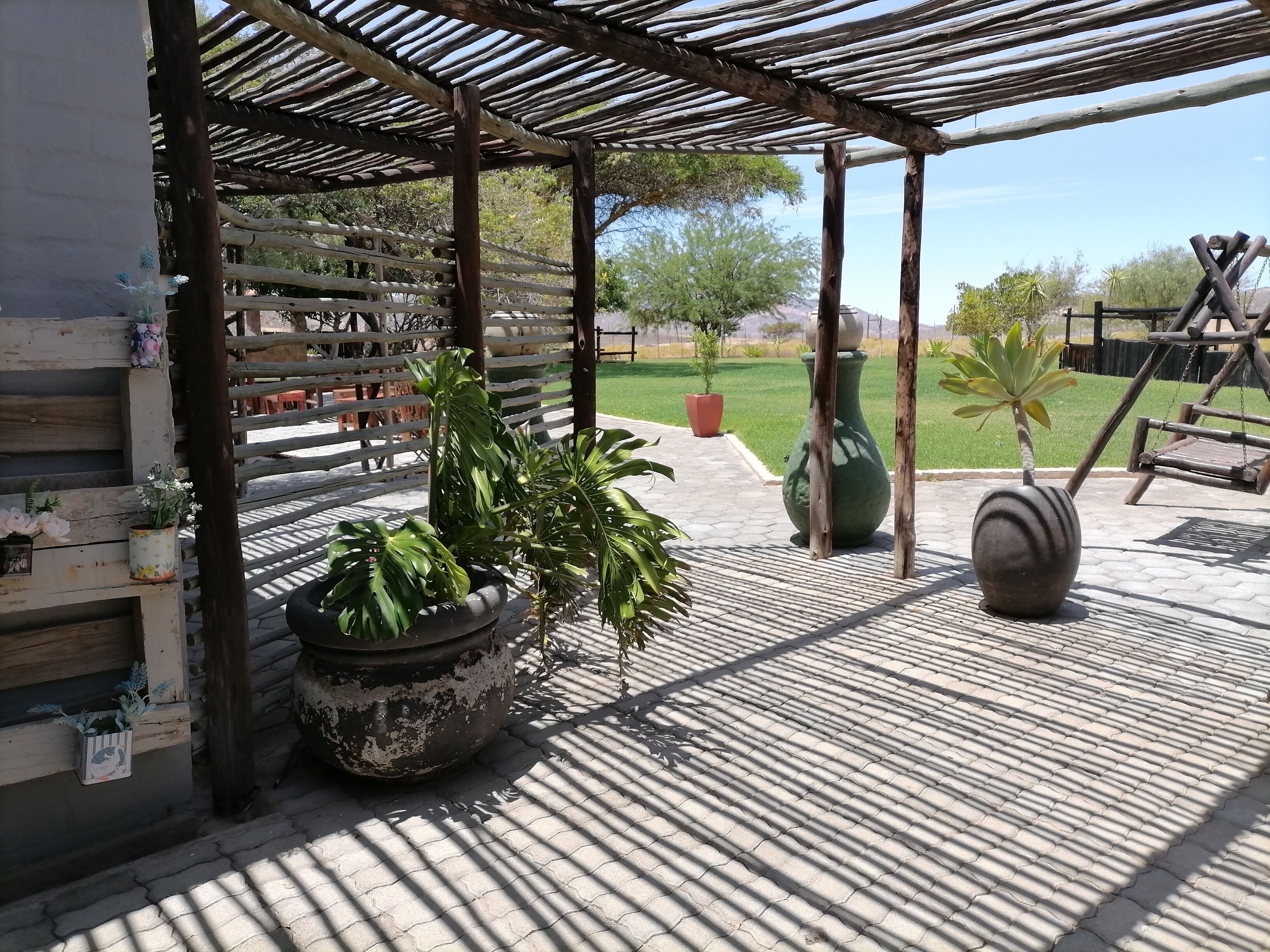 Uma área externa coberta com pérgola de madeira, plantas em vasos, balanço e vista para um campo gramado.