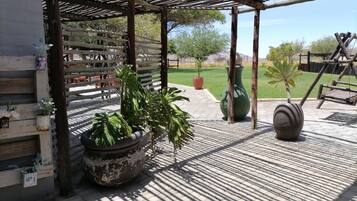Uma área externa coberta com pérgola de madeira, plantas em vasos, balanço e vista para um campo gramado.