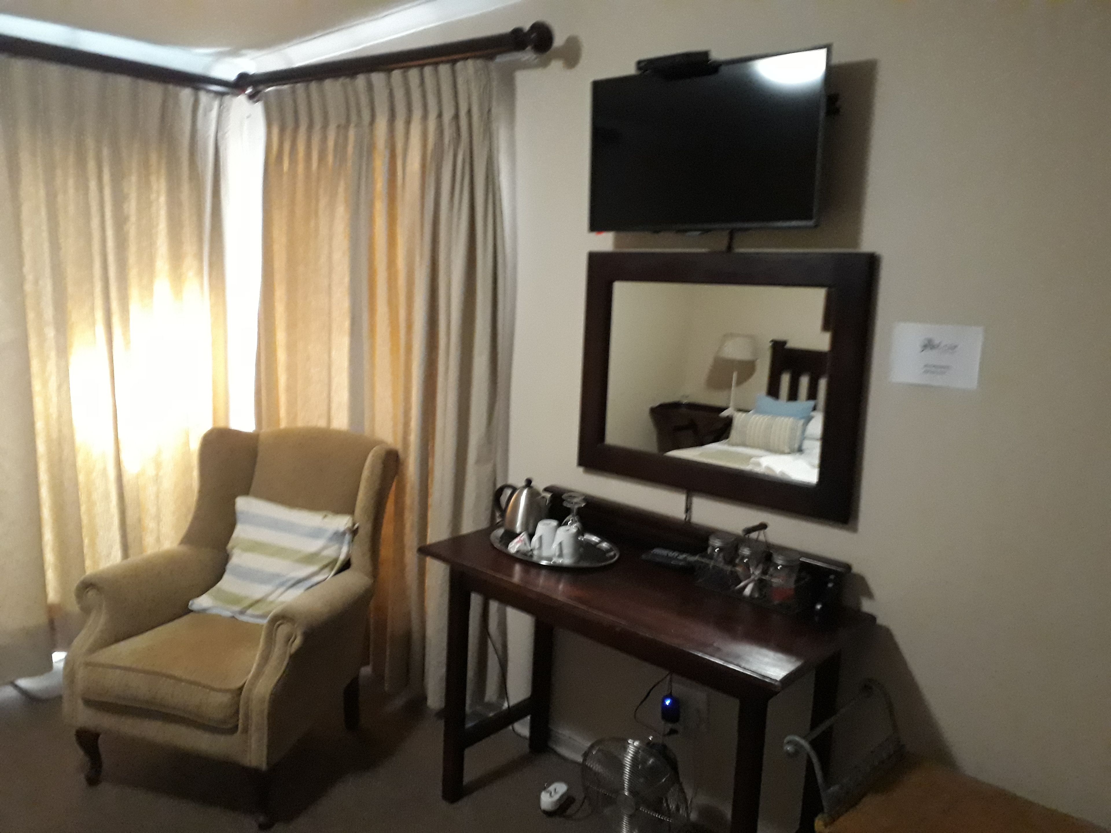 Quarto de hotel com uma poltrona, uma mesinha com chaleira e bandeja, uma TV de tela plana e um espelho refletindo o interior do quarto.