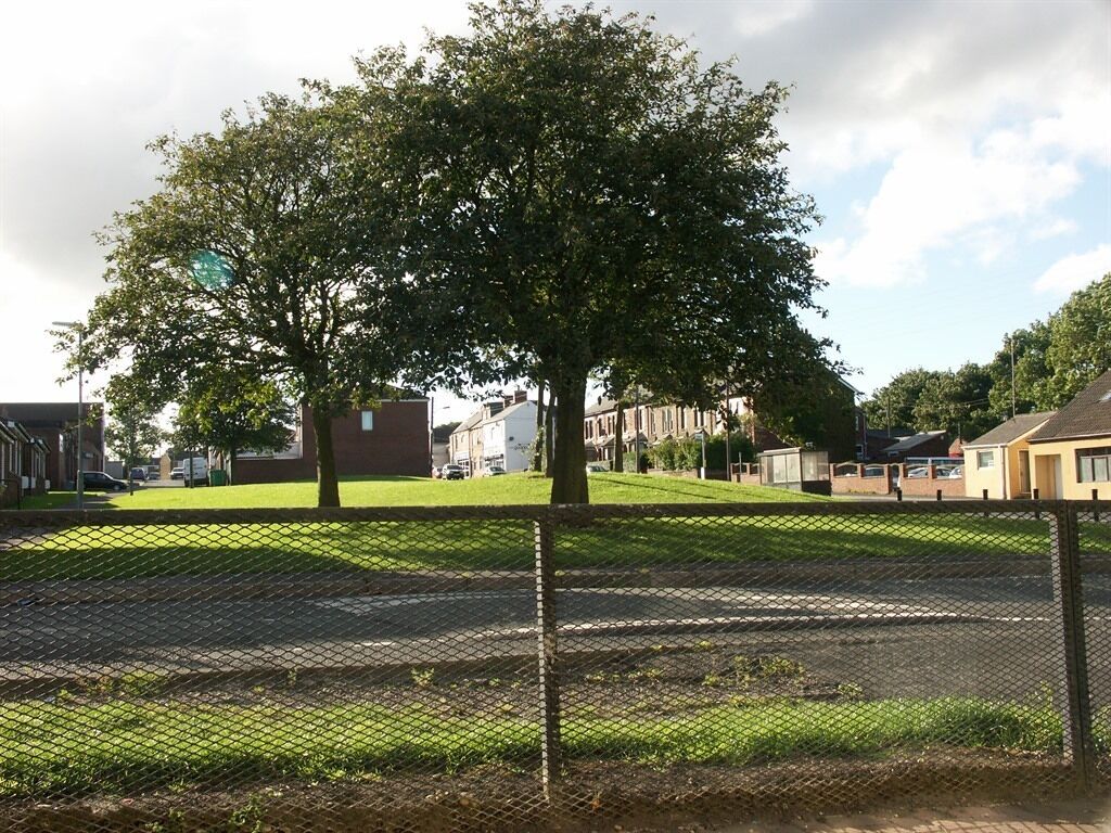 Área externa cercada com uma árvore, um campo gramado e edifícios residenciais ao fundo.
