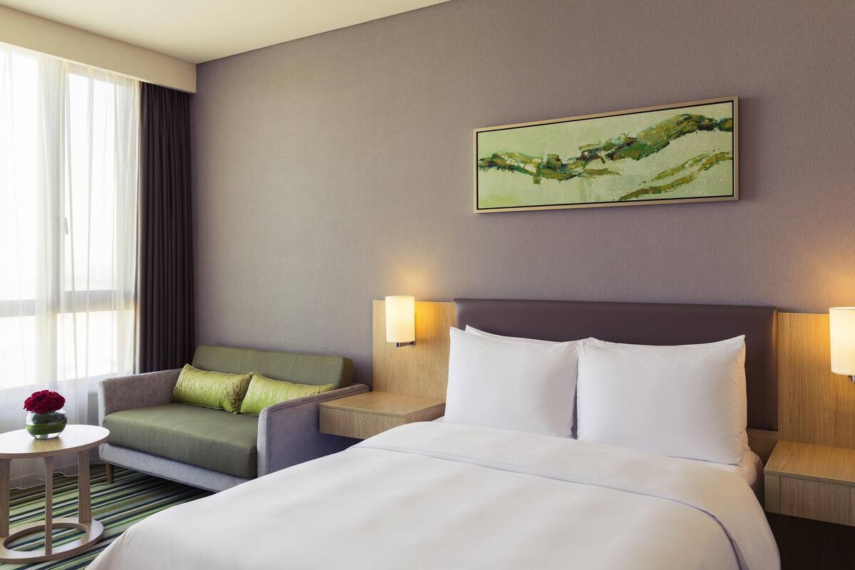 Quarto de hotel com uma cama, um sofá verde, uma mesa pequena com um vaso de flores e um quadro na parede.