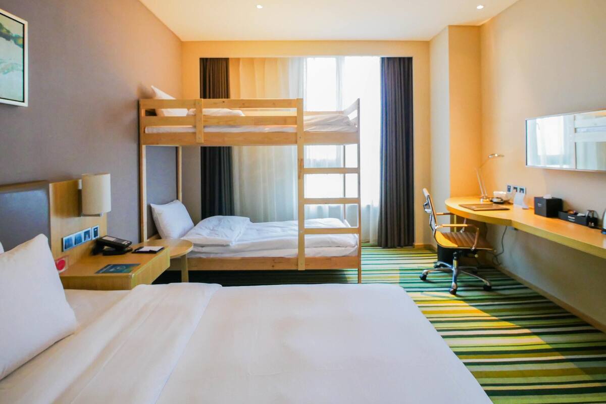 Quarto de hotel com uma cama beliche, uma mesa com cadeira, uma luminária, um telefone e uma janela com cortinas.