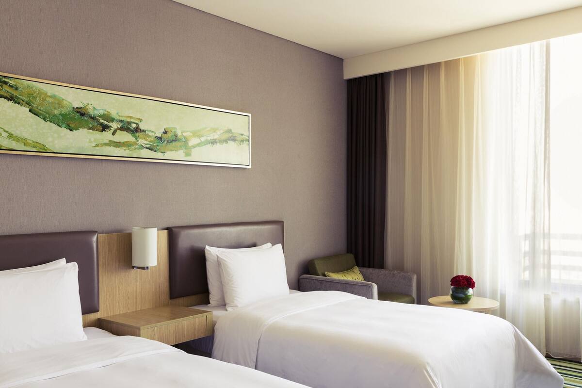 Quarto de hotel com duas camas, uma mesa de cabeceira, abajur, cadeira, quadro e janela com cortinas.