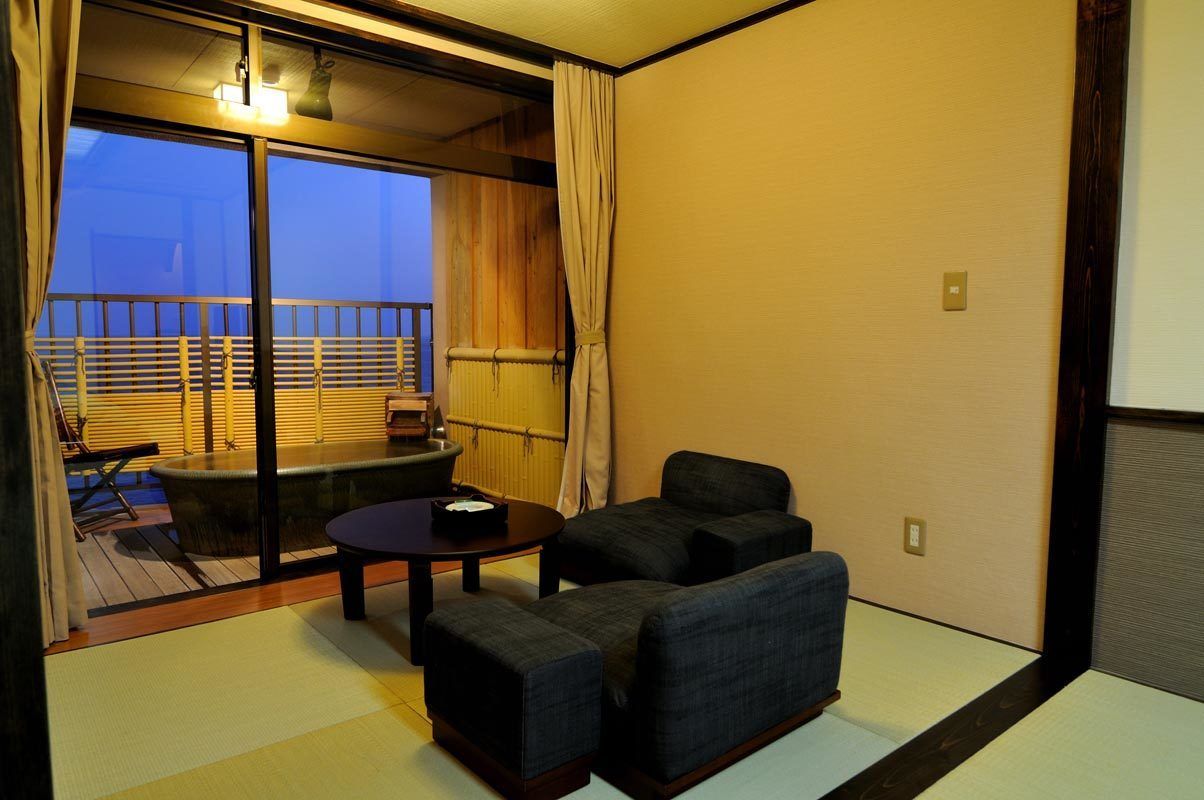 Quarto com piso de tatame, mesa baixa com almofadas, sofá e porta de vidro deslizante que dá para uma varanda com guarda-corpo de madeira.