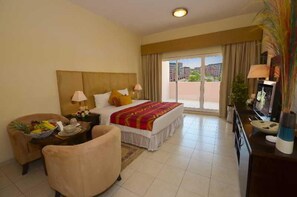 Quarto de hotel com cama, uma cadeira, uma mesinha com uma cesta de frutas, uma luminária e vista para prédios pela janela.