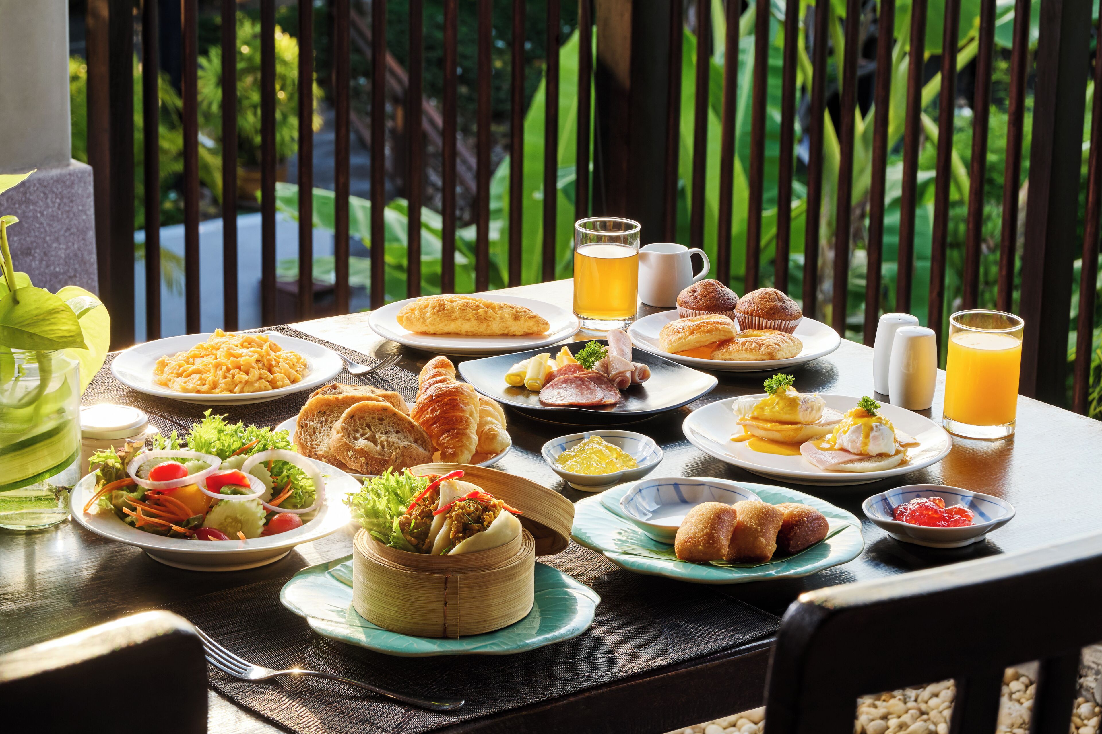 Mesa posta com diversos pratos de café da manhã, incluindo ovos, doces e uma salada, com vista para o verde ao fundo.