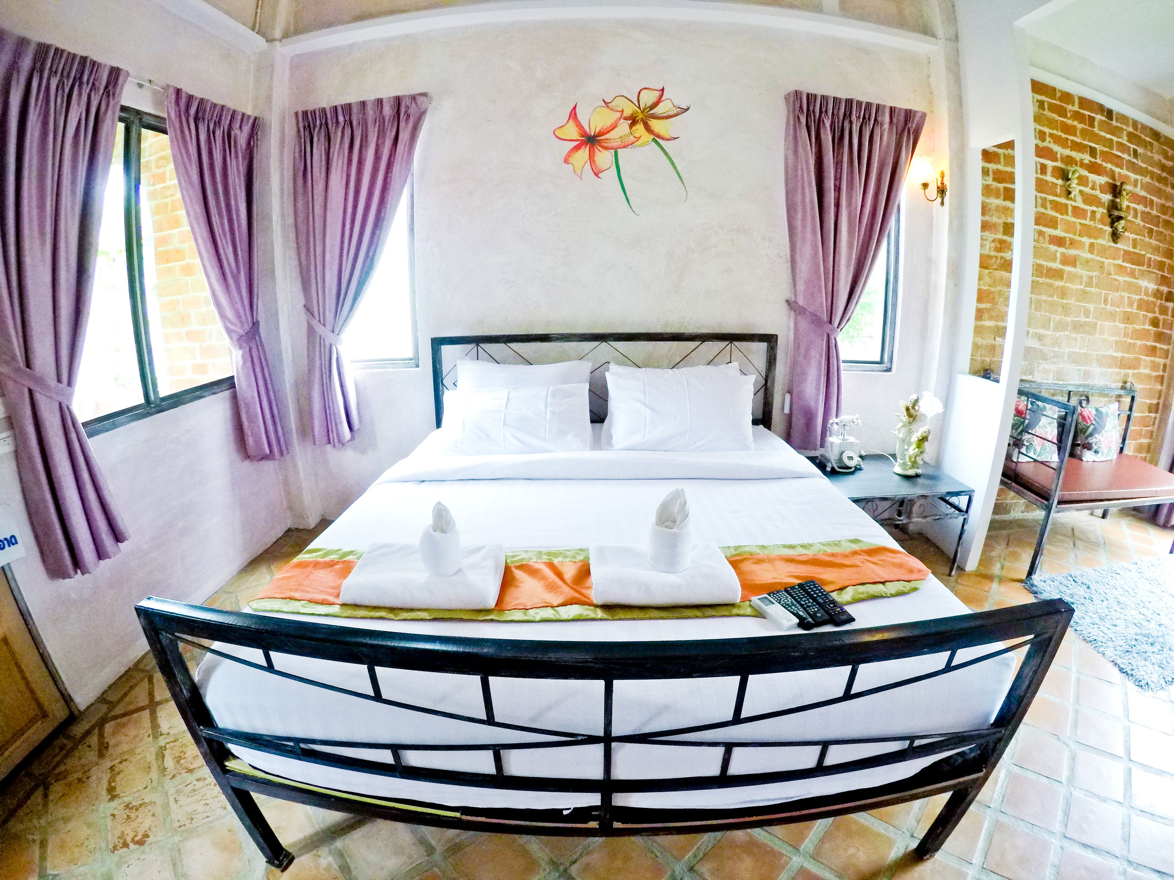 Quarto de hotel com cama, cortinas roxas, parede de tijolos e decoração floral.