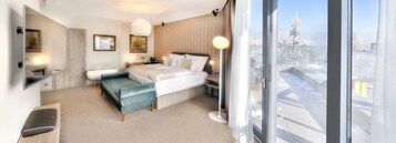 Quarto de hotel moderno com cama, um banquinho, mesa de cabeceira e uma janela grande com vista para a cidade.