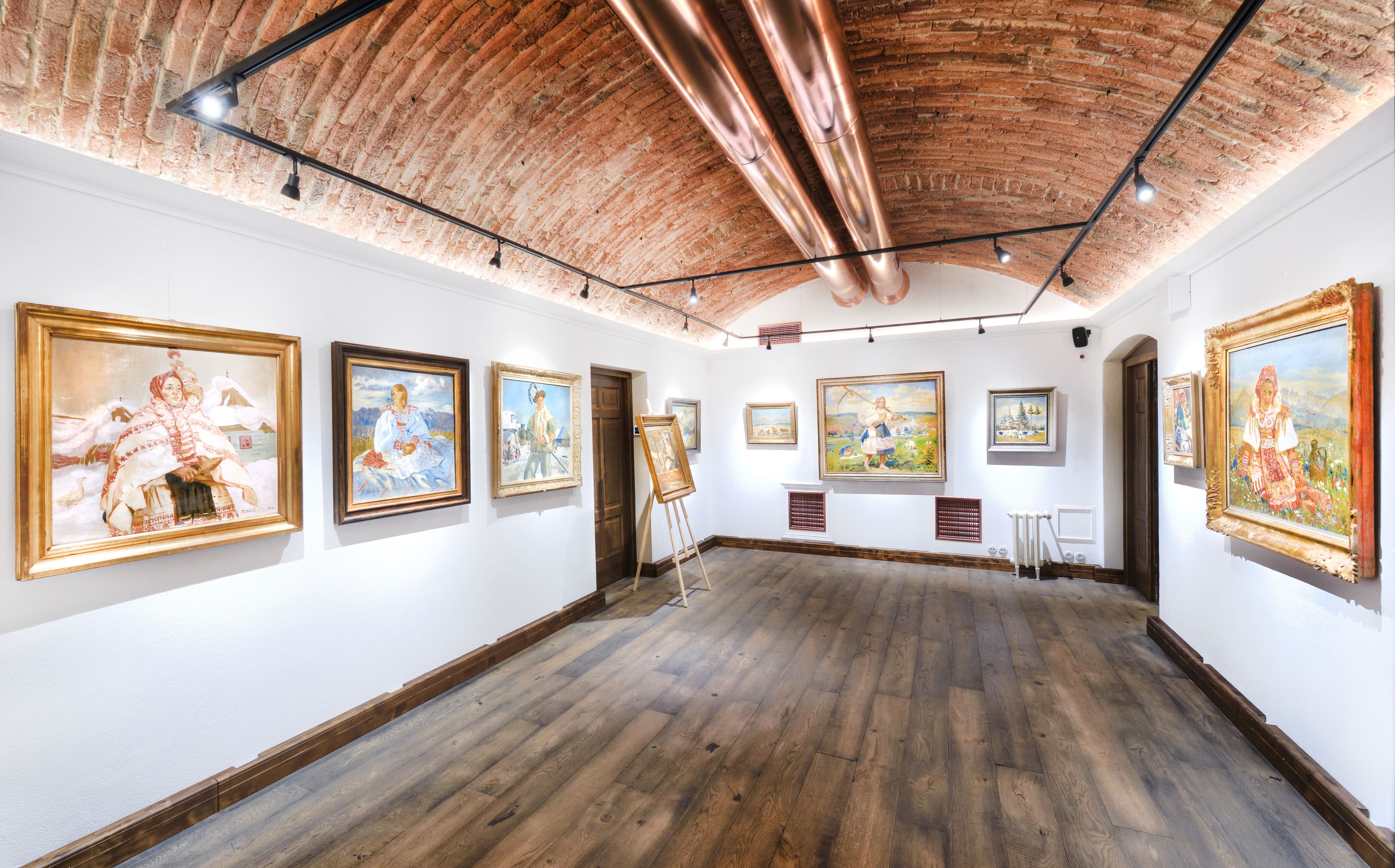 Uma galeria de arte com pisos de madeira e teto de tijolos, exibindo diversas pinturas emolduradas.