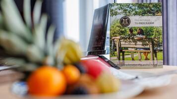 Programa de televisão exibindo um casal sentado em um banco em um parque, um laptop e algumas frutas sobre uma mesa.