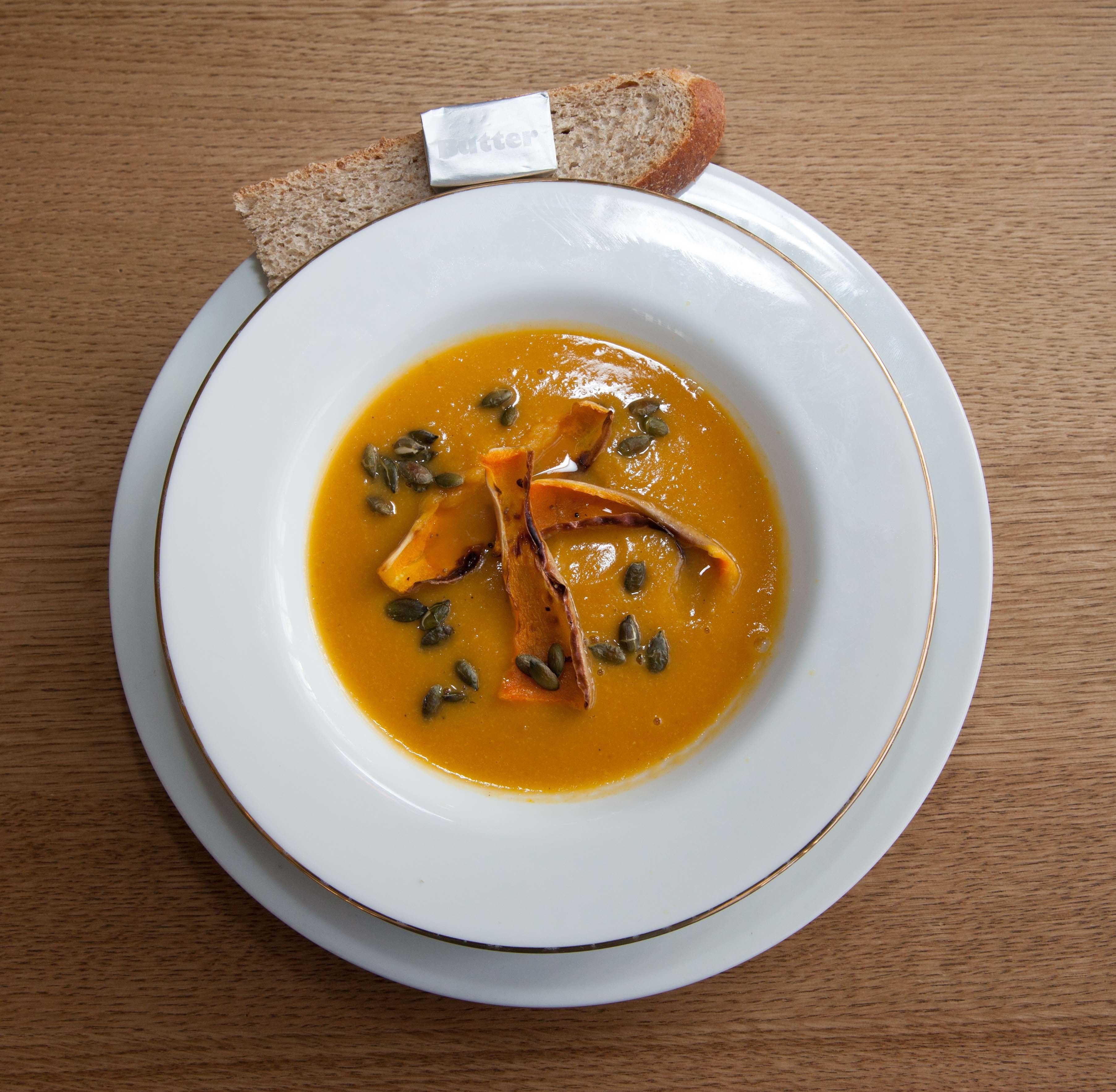 Um prato branco com sopa adornada com sementes de abóbora e bacon, acompanhado de uma fatia de pão sobre uma mesa de madeira.