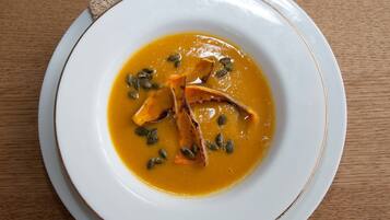 Um prato branco com sopa adornada com sementes de abóbora e bacon, acompanhado de uma fatia de pão sobre uma mesa de madeira.