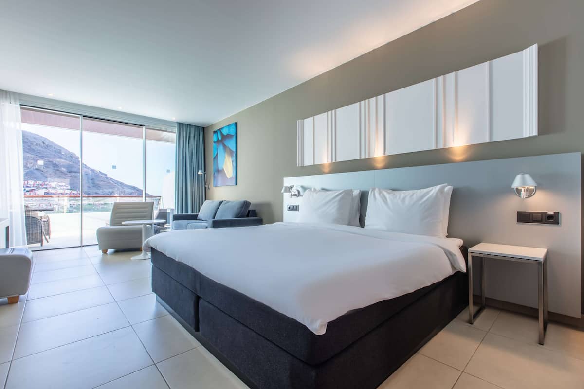 Quarto de hotel moderno com uma cama grande, uma mesa pequena, uma cadeira e vista da cidade através de janelas amplas.