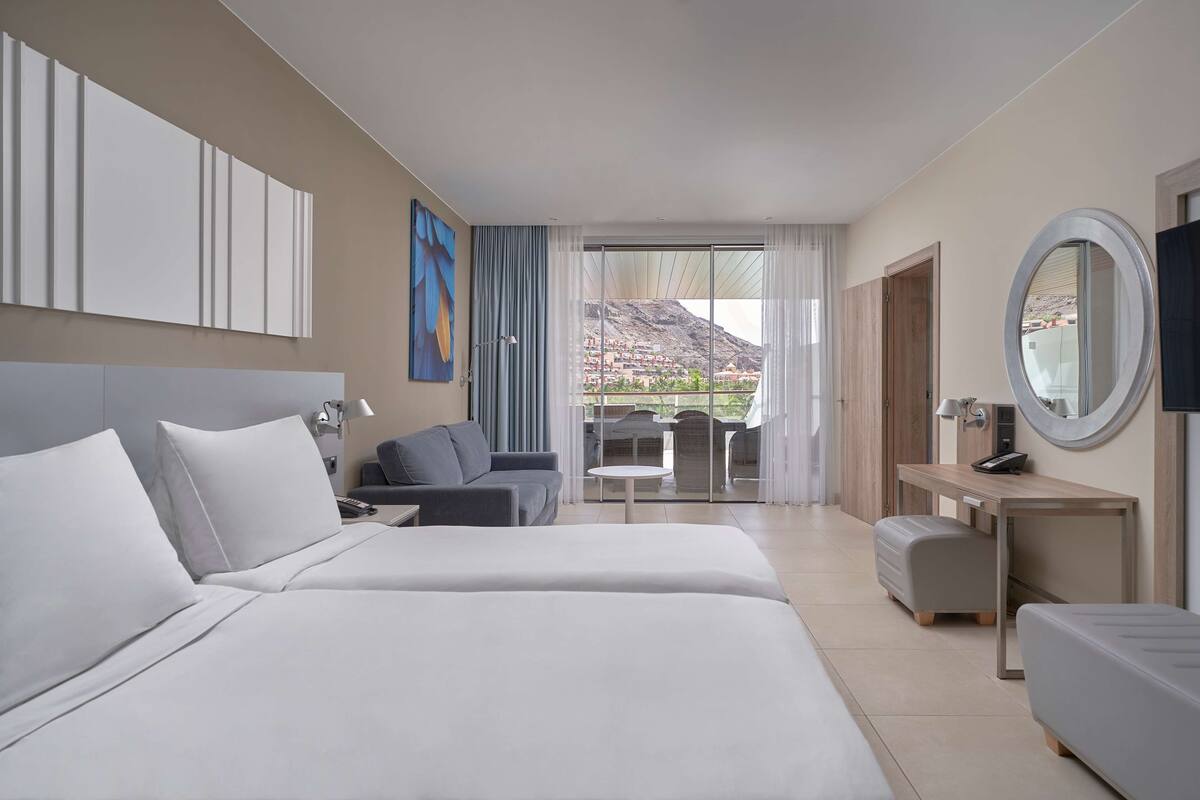 Quarto de hotel com duas camas, uma escrivaninha, uma cadeira, televisão e varanda com vista para as montanhas.