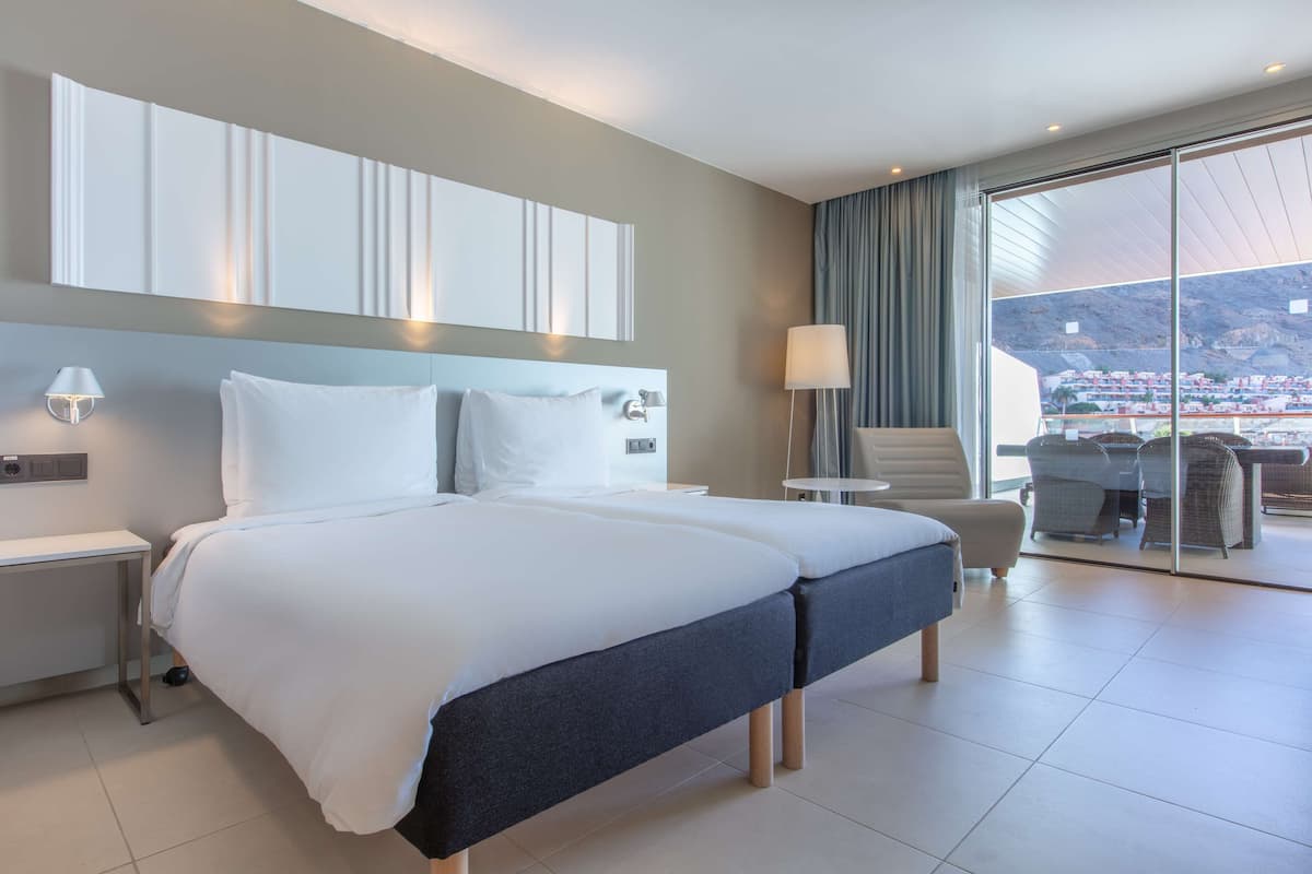 Quarto de hotel com uma cama grande, uma mesa, uma cadeira e vista para uma paisagem montanhosa.