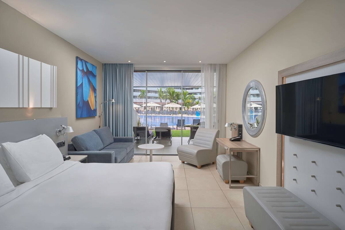 Quarto de hotel com cama, sofá, televisão, mesa pequena e varanda com área de estar.