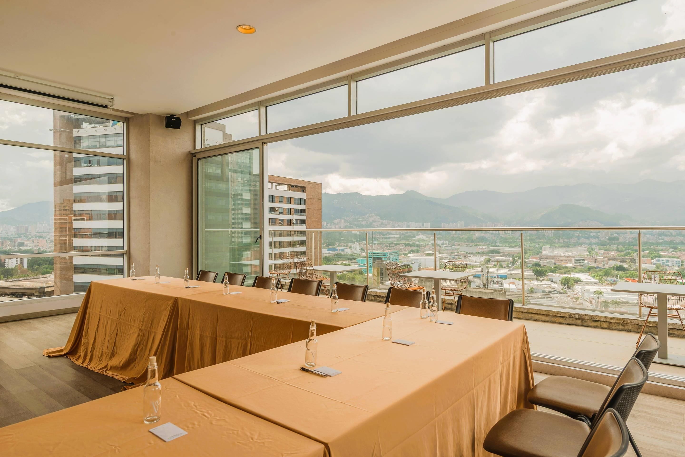 Uma sala de conferências com mesas preparadas para reuniões, grandes janelas com vista para a paisagem urbana e um edifício moderno ao fundo.