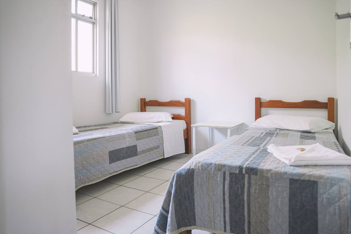 Quarto pequeno e limpo com duas camas de solteiro, uma mesinha e uma janela.