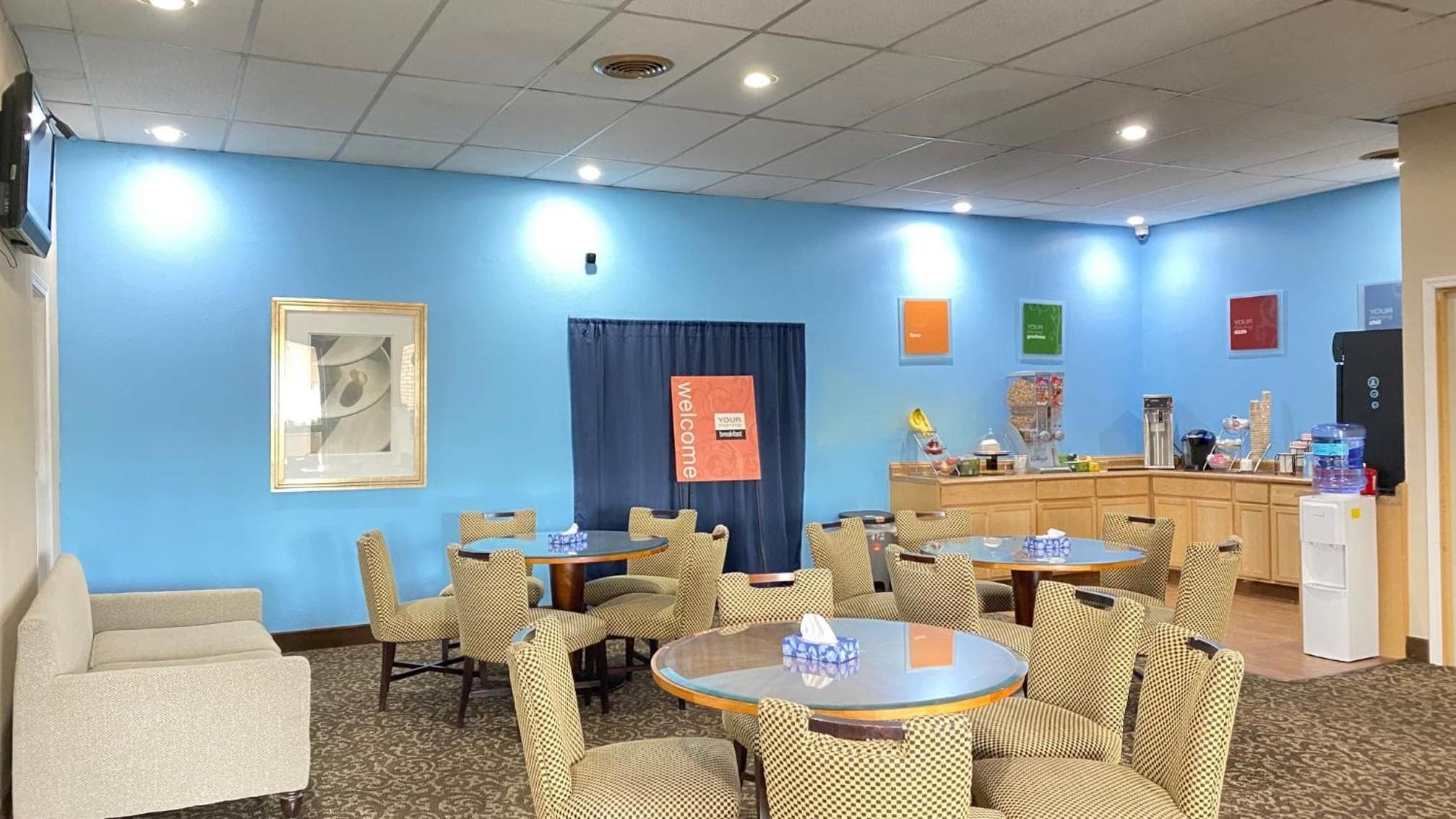 Um saguão de hotel com paredes azuis, balcão de recepção e áreas de estar com mesas redondas e cadeiras estampadas.