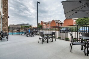 Uma área de pátio externa com mesas e cadeiras, piscina e estacionamento ao fundo.