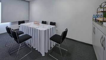 Uma sala de conferência com uma mesa branca, cadeiras pretas, uma tela de projetor e um buffet com comida e bebidas.