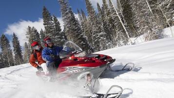 Uma moto de neve com dois passageiros, cercada por neve e árvores.