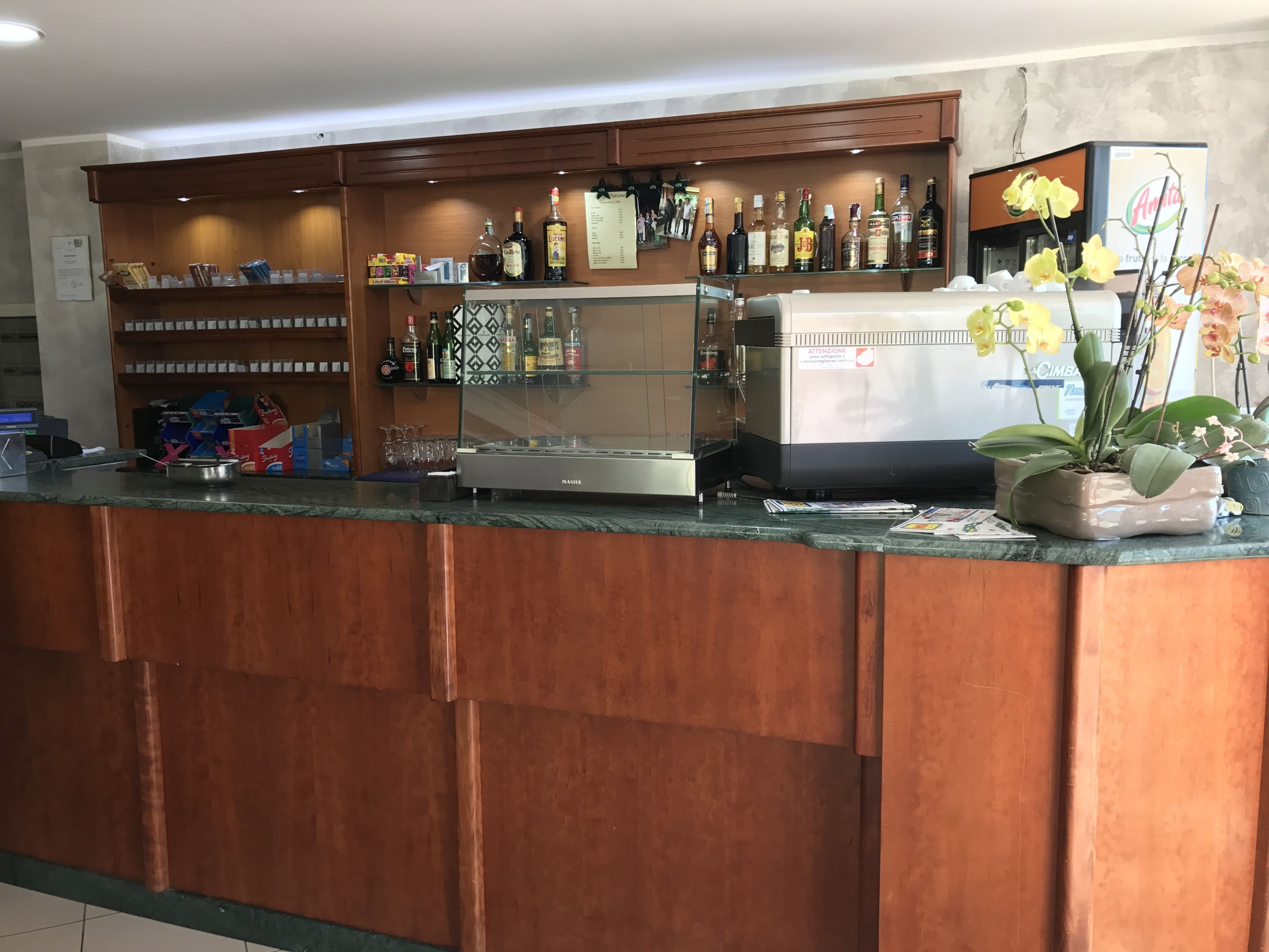 Uma área de bar com máquina de café, prateleiras de garrafas de bebidas e uma orquídea em vaso.