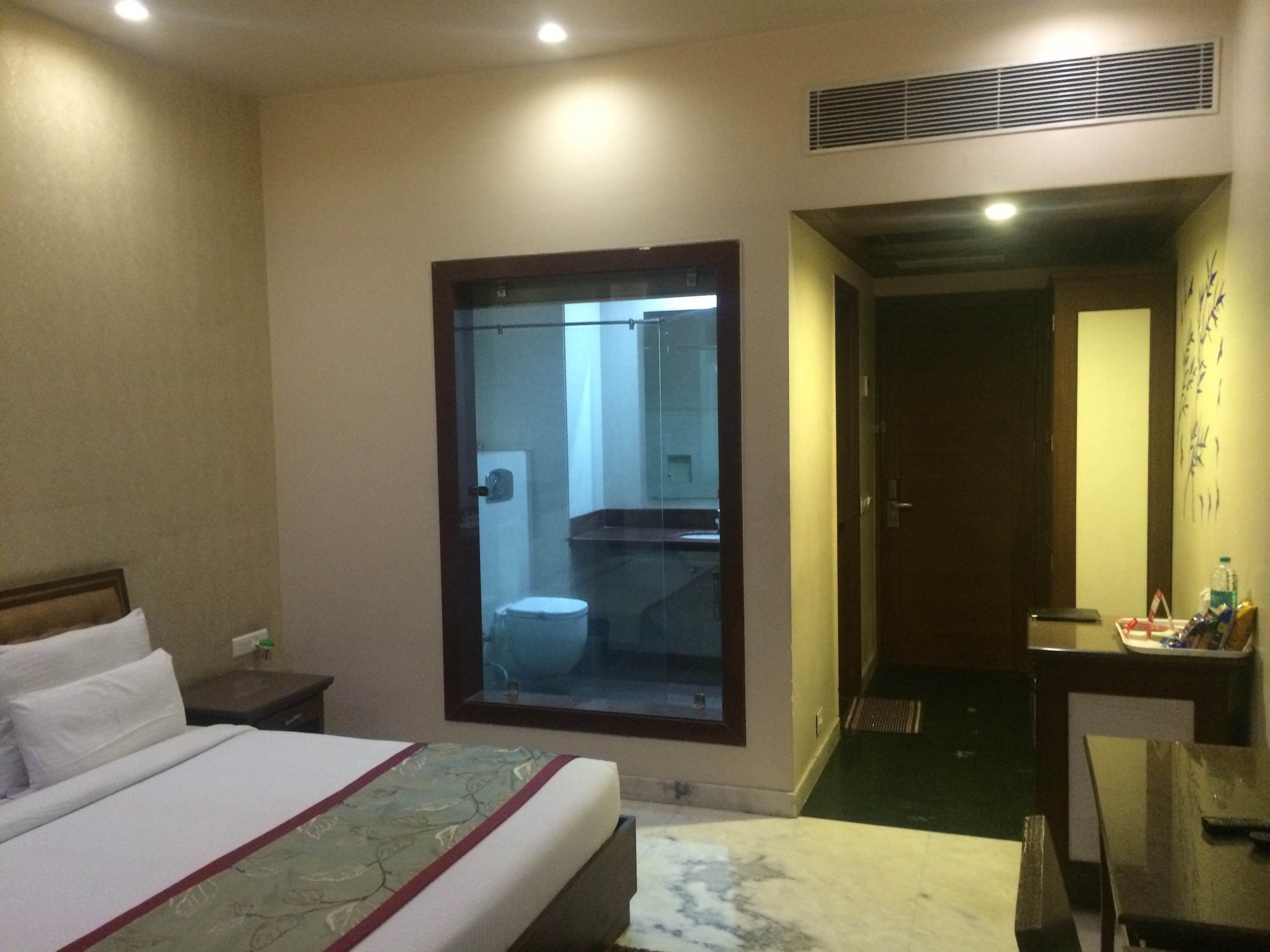 Quarto de hotel com cama, escrivaninha, banheiro visível através de uma porta de vidro e uma pequena área de cozinha.