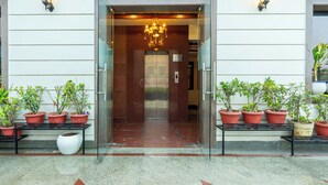 Entrada de hotel com porta de vidro, um candelabro, plantas em vasos e um banco.