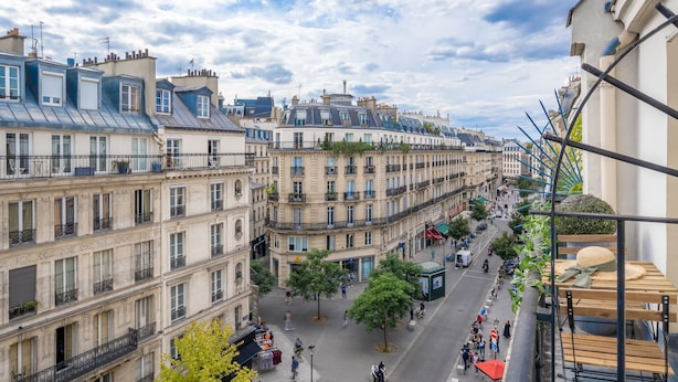 Vista da rua com edifícios altos, calçada arborizada e céu claro.