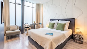 Quarto de hotel moderno com uma cama grande, uma escrivaninha pequena, uma cadeira e vista da cidade através de grandes janelas.