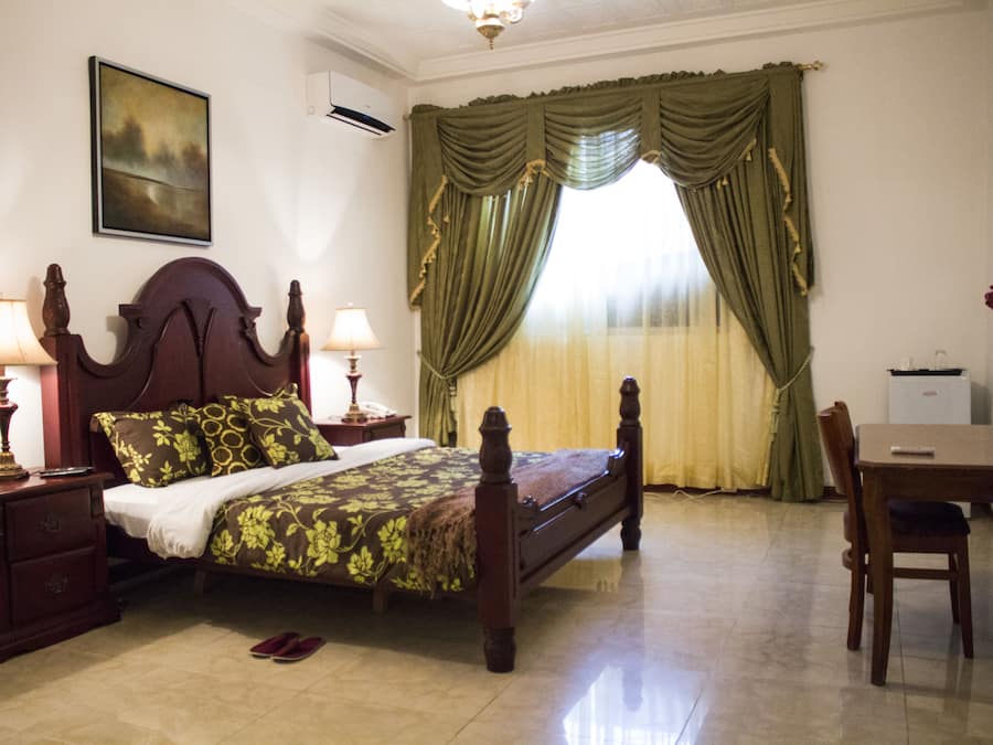 Quarto com cama de madeira, duas mesas de cabeceira com luminárias, uma mesinha com uma cadeira e uma janela com cortinas verdes.