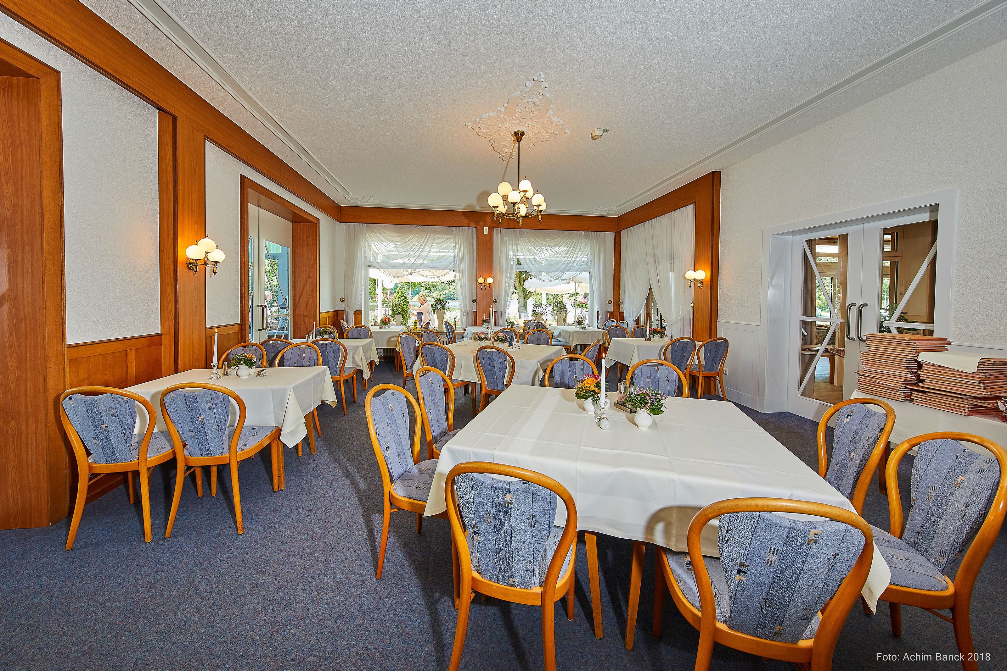 Uma grande sala de jantar com várias mesas e cadeiras, um lustre e amplas janelas com cortinas.
