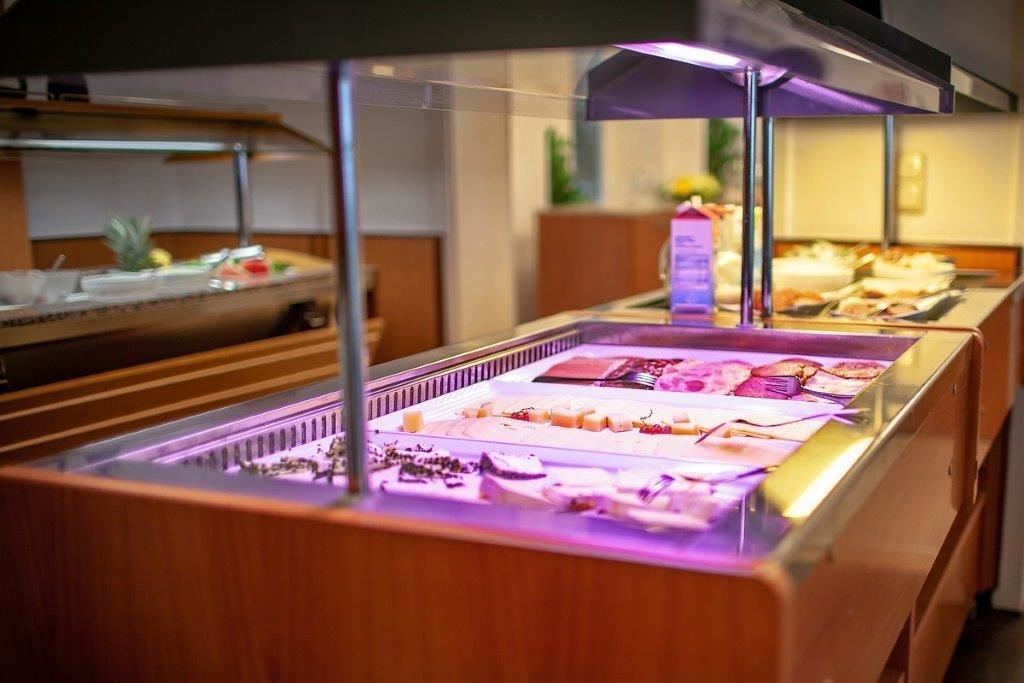 Uma área de buffet com diversos alimentos expostos em vitrine de vidro, iluminada por luzes roxas.