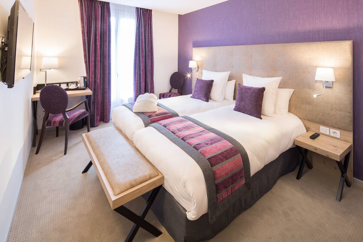 Quarto de hotel com duas camas, uma escrivaninha, uma cadeira e um banco.