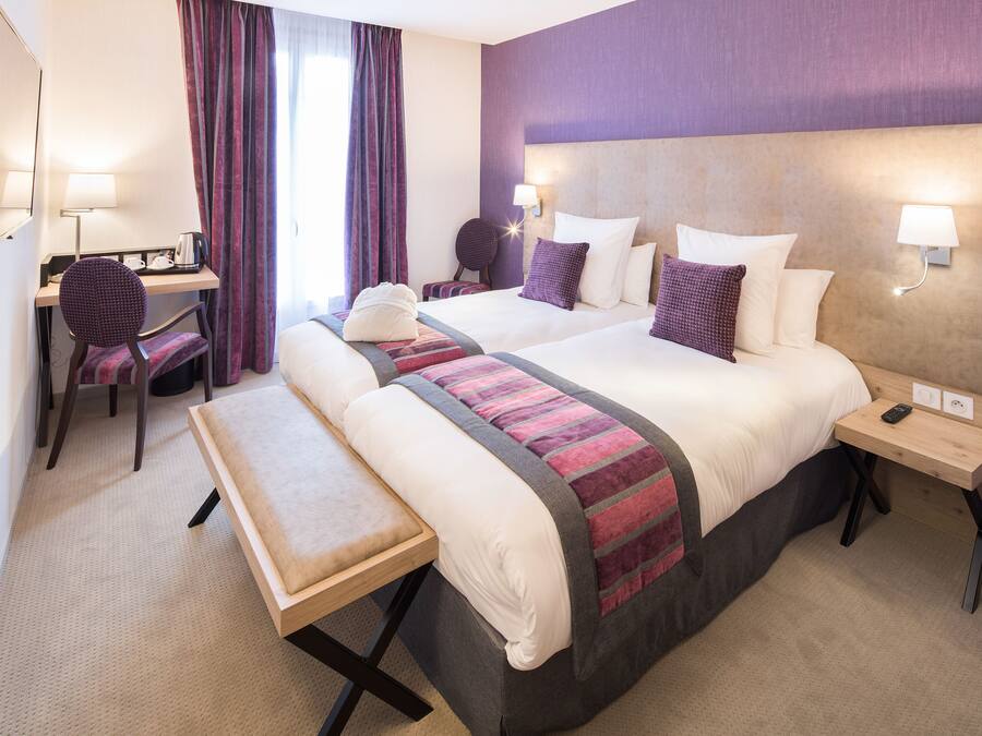 Quarto de hotel com duas camas, uma escrivaninha, uma cadeira e um banco.