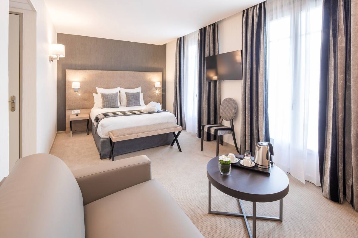 Quarto de hotel moderno com uma cama grande, mesas de cabeceira, um sofá, uma pequena mesa com uma chaleira e uma TV fixada na parede.