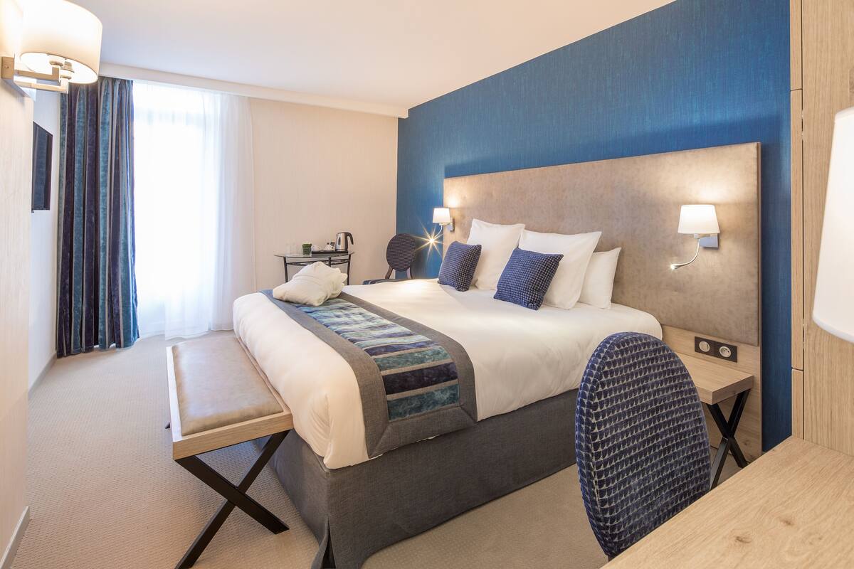 Quarto de hotel com uma cama grande, uma escrivaninha, uma cadeira, um banco, uma luminária de mesa, uma mesa de cabeceira e uma janela com cortinas.