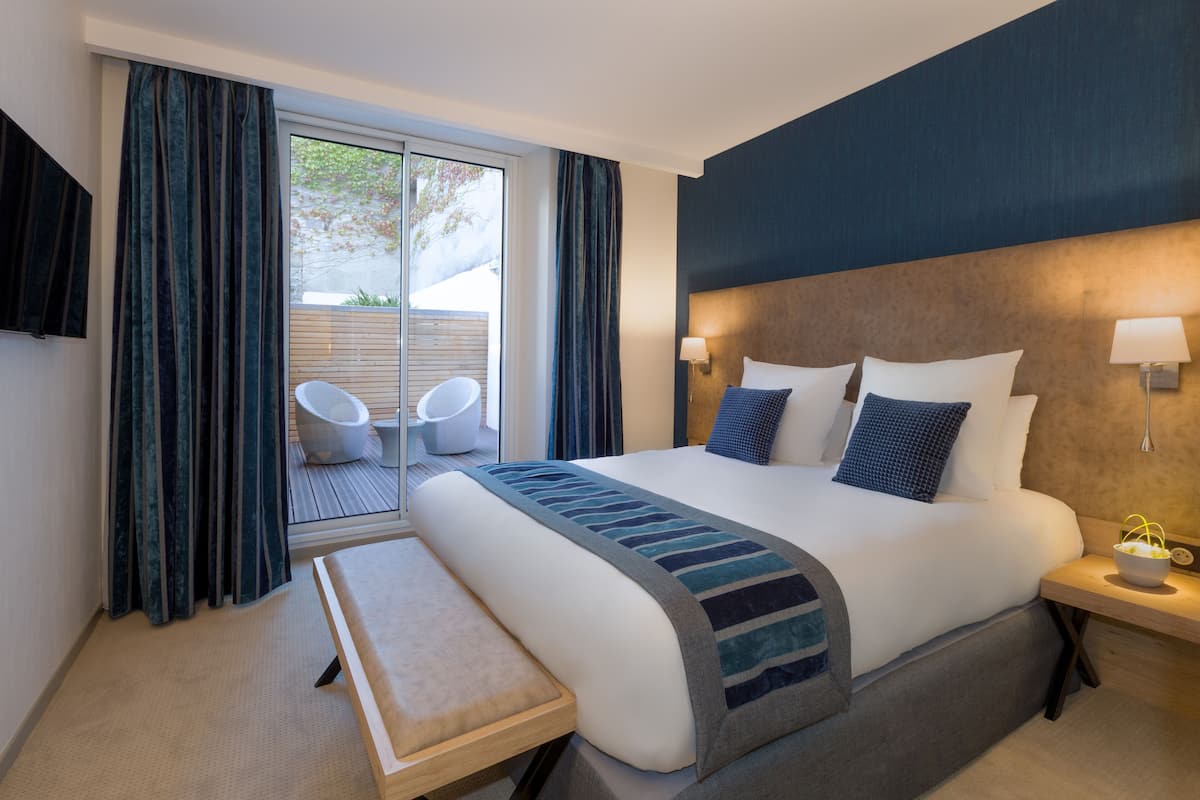 Quarto de hotel com uma cama grande, uma mesa de cabeceira com luminária, um banco e vista para o exterior através de uma porta de correr.