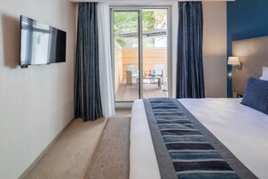 Quarto de hotel com uma cama grande, televisão de tela plana presa à parede, porta de vidro deslizante para uma área de estar externa e mesa de cabeceira com abajur.