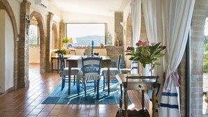Uma sala de jantar com vista, com uma mesa posta, cadeiras, um tapete azul e um vaso com flores.