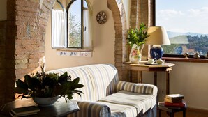 Um quarto aconchegante com um sofá listrado, uma mesa com um vaso e livros, e vista para o campo através de uma janela.