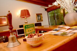 Uma área de recepção de hotel com um balcão de madeira, um recipiente com lanches, uma placa com a inscrição 'Albergo', uma luminária e um vaso com flores.