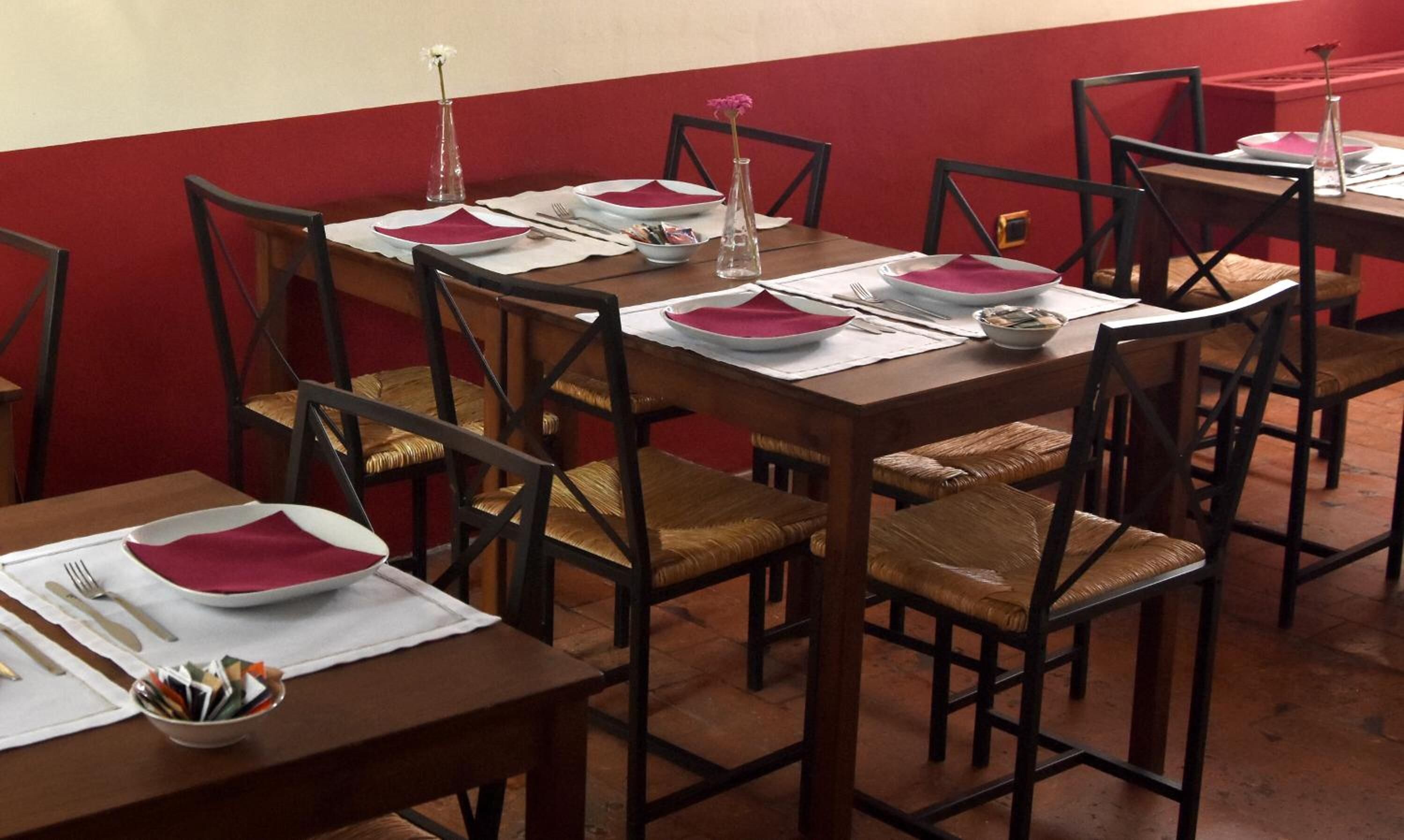 Uma sala de jantar com mesas e cadeiras de madeira, cada conjunto com jogos americanos brancos, guardanapos vermelhos e vasos de vidro transparente com flores.