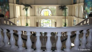 Um interior imponente com uma janela central de vitrais, um corrimão branco e palmeiras decorativas.