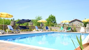 Piscina com espreguiçadeiras e guarda-sóis, cercada por muros de pedra e vegetação.