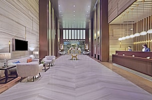 Um hall amplo com design moderno, apresentando uma área de estar central com sofás e poltronas, um balcão de recepção e um lustre grande.